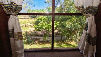 Quarto com uma janela grande, cortinas com padrões listrados e vista para um jardim com árvores e uma estrutura de pedra.