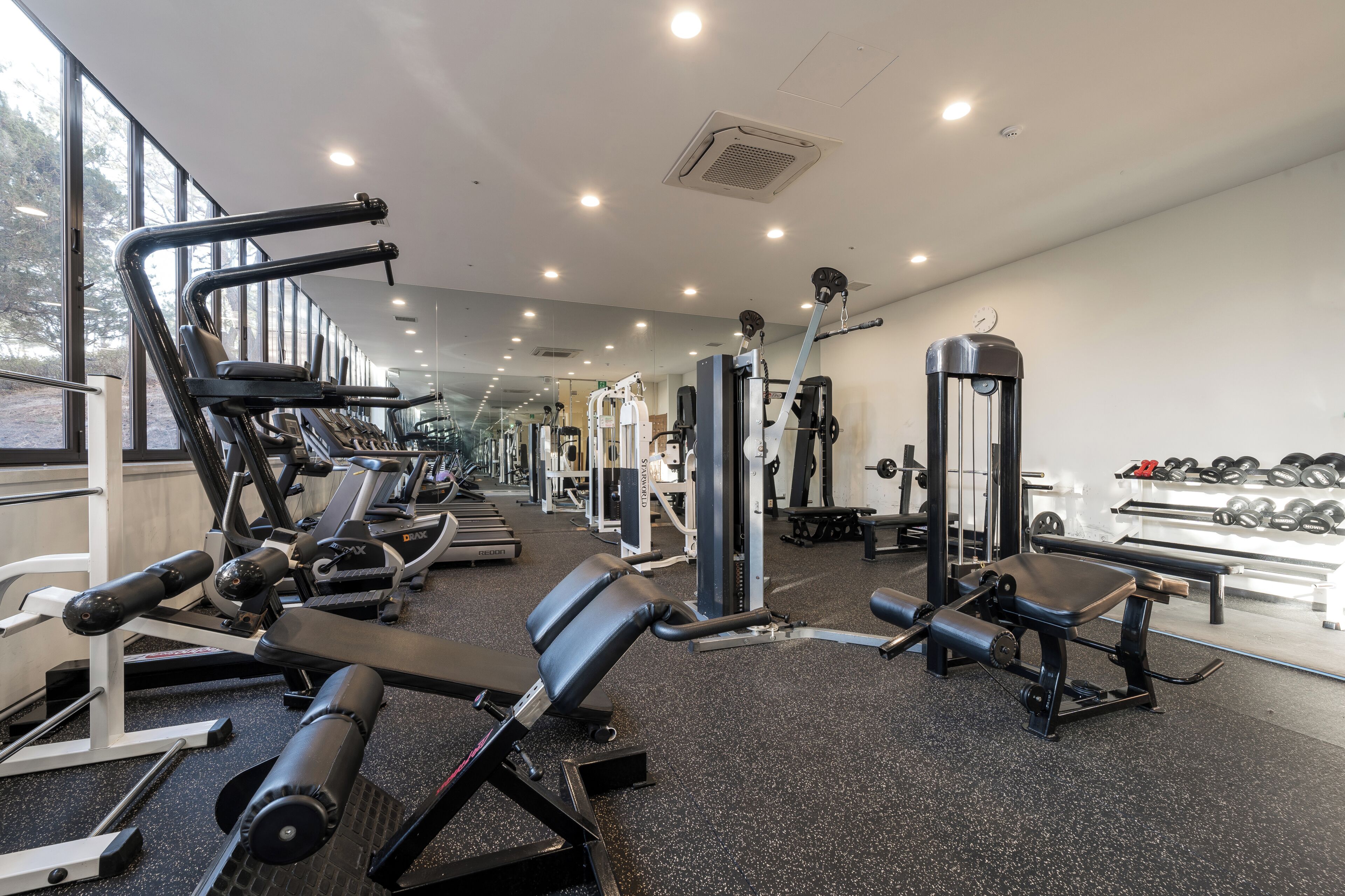Academia com diversos equipamentos de exercício, amplas janelas e um espelho grande.
