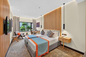 Quarto de hotel com duas camas, uma escrivaninha, uma cadeira, uma televisão e vista para o verde lá fora.