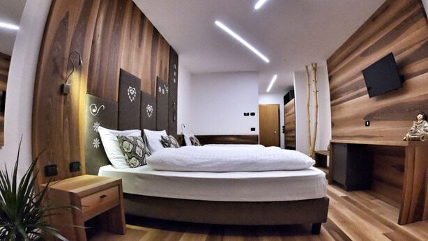 Um quarto moderno com cabeceira de madeira, uma cama grande com várias almofadas, uma mesinha de cabeceira e uma TV fixada na parede.