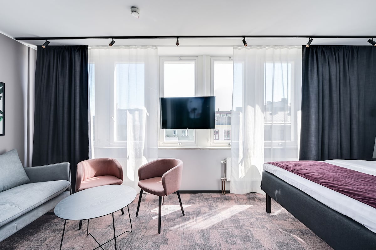 Quarto de hotel com cama, sofá, poltrona, mesa pequena e televisão.