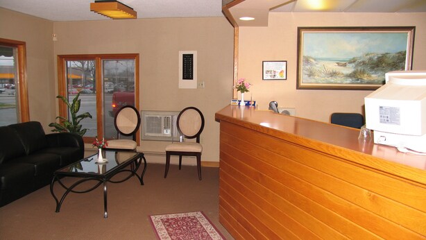 Uma área de recepção com uma mesa de madeira, duas cadeiras, um sofá preto, uma mesa de vidro, um vaso com flores, um quadro na parede e uma janela com vista para carros do lado de fora.