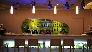 Uma área de bar com uma estante iluminada em verde exibindo diversas garrafas, um balcão e cadeiras de madeira.