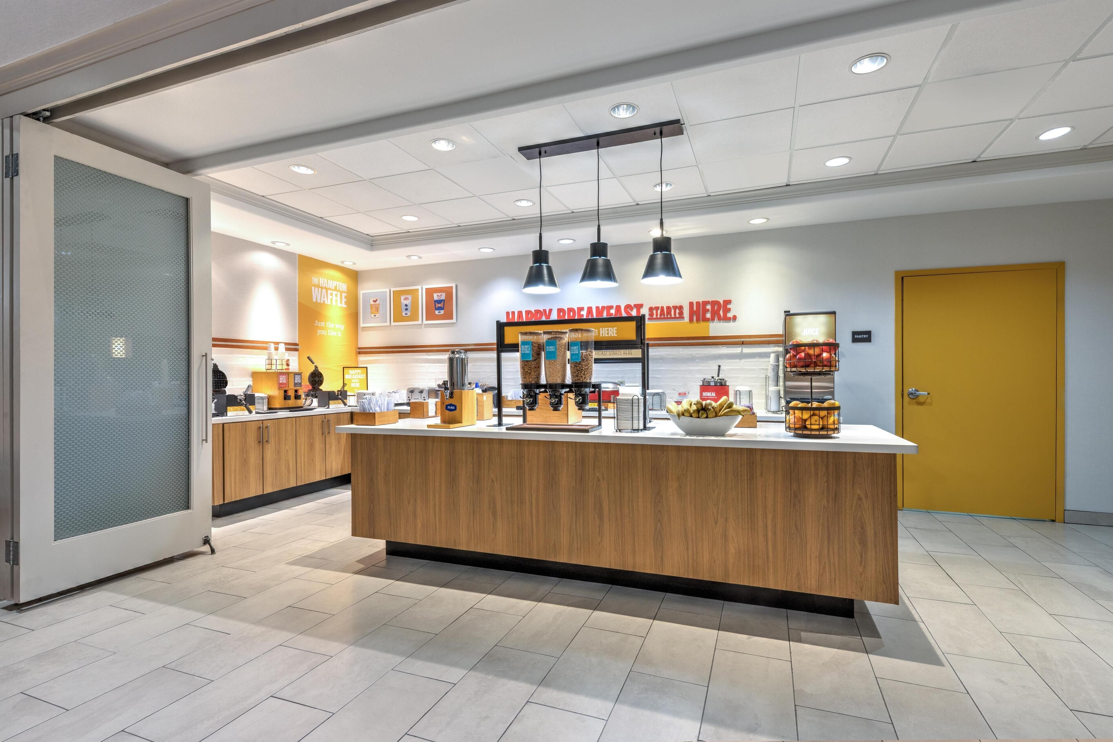 Um café moderno com balcão de madeira, estação de waffles e um letreiro que diz 'HAPPY BREAKFAST START HERE'.