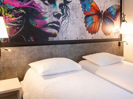 Quarto de hotel com uma grande pintura abstrata na parede, uma cama com roupa de cama branca e uma mesa de cabeceira com abajur.