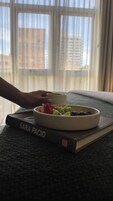 Uma tigela de frutas sobre um livro intitulado "SARA FACIO", perto de uma janela com persianas.