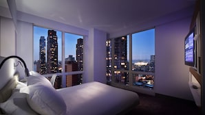 Quarto com cama grande, mesa de cabeceira, televisão e vista para a cidade ao entardecer.