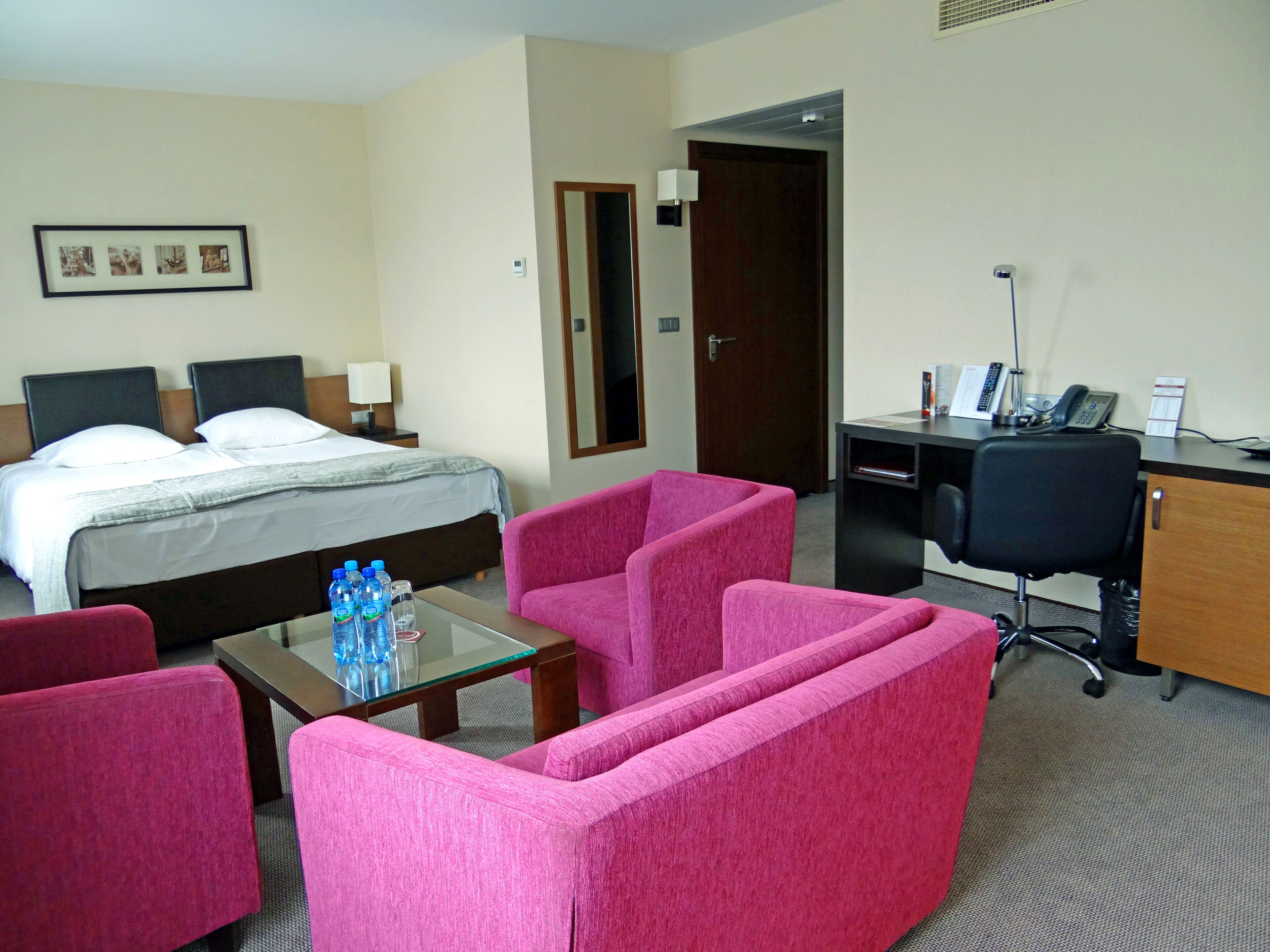 Quarto de hotel com cama, escrivaninha, cadeira e uma pequena mesa com garrafas de água.