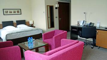 Quarto de hotel com cama, escrivaninha, cadeira e uma pequena mesa com garrafas de água.