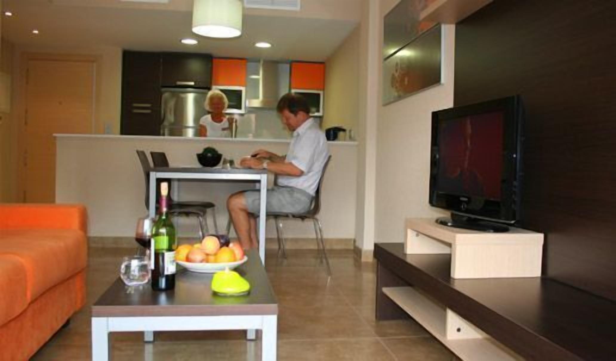 Uma sala de estar moderna com uma mesa de jantar, um sofá, uma TV e uma cozinha ao fundo.