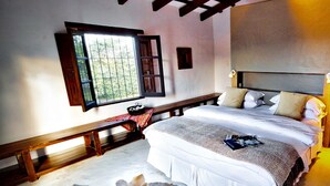 Quarto com cama grande, um banquinho, tapete e janela com vista para área verde.