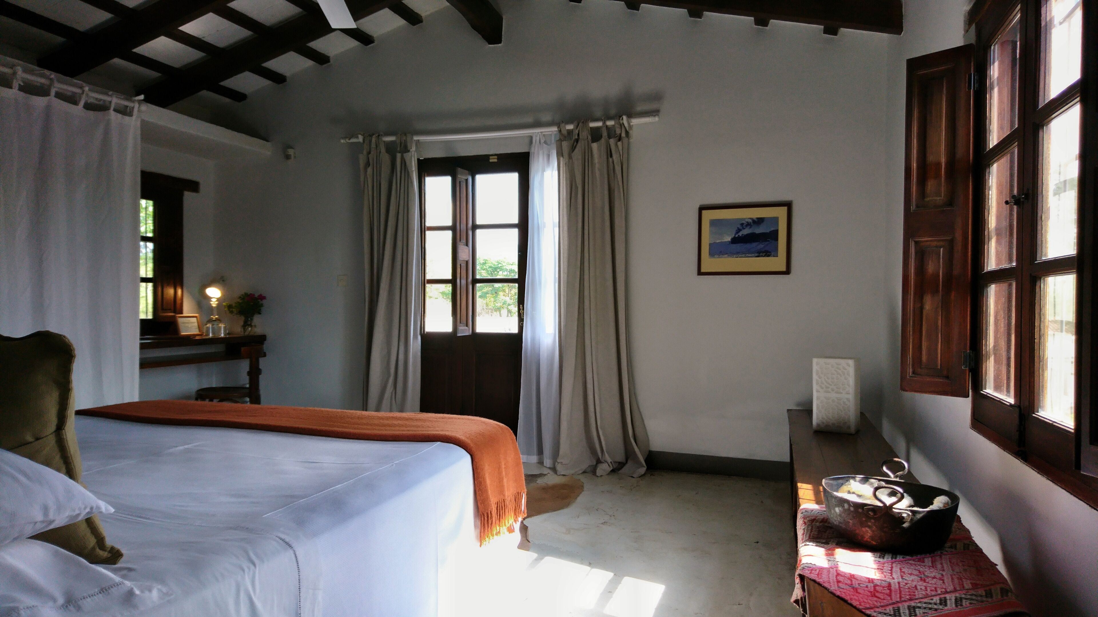 Quarto com cama, escrivaninha, cadeira, janela com cortinas e um quadro na parede.