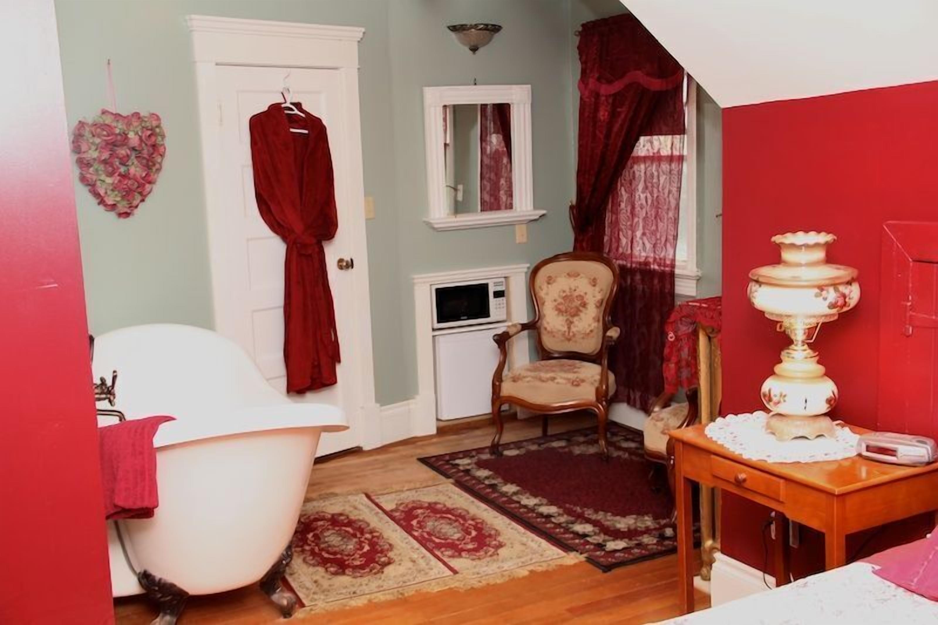 Um quarto com uma banheira branca, uma cadeira de madeira, uma mesa pequena com um abajur e uma parede vermelha.
