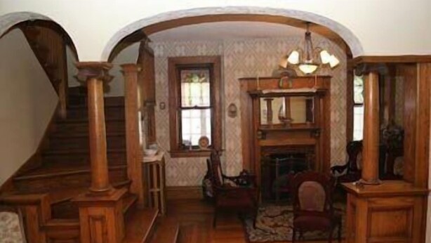 Uma escadaria imponente com guarda-corpos de madeira e um candelabro, que leva a uma sala de estar com lareira e uma janela com cortinas.