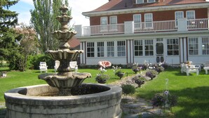 Uma casa grande de dois andares com um jardim bem cuidado, incluindo uma fonte e diversos vasos de plantas.