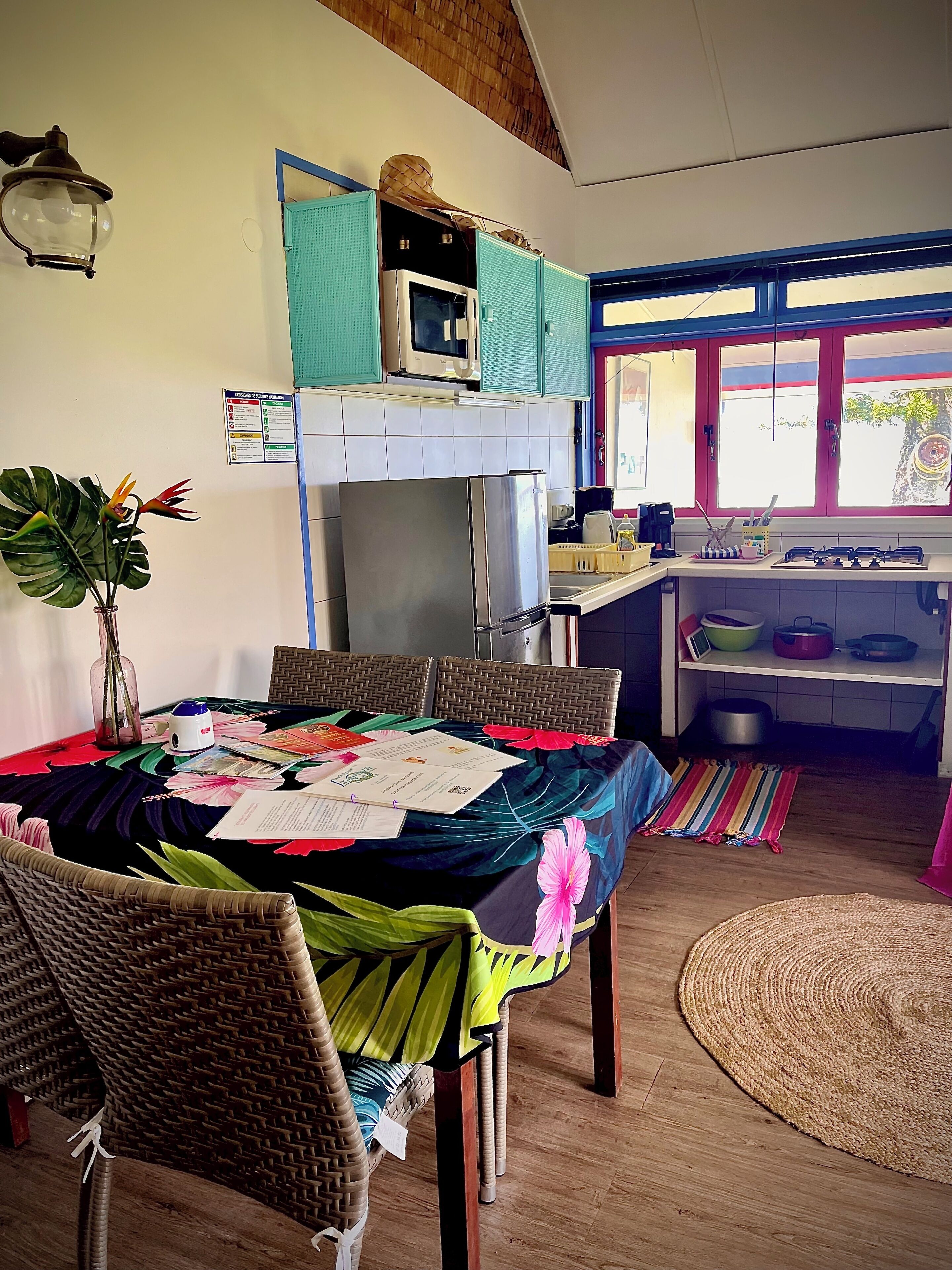 Uma sala de jantar com uma mesa posta para quatro pessoas, cercada por cadeiras de vime, e uma cozinha com micro-ondas, fogão e pia.