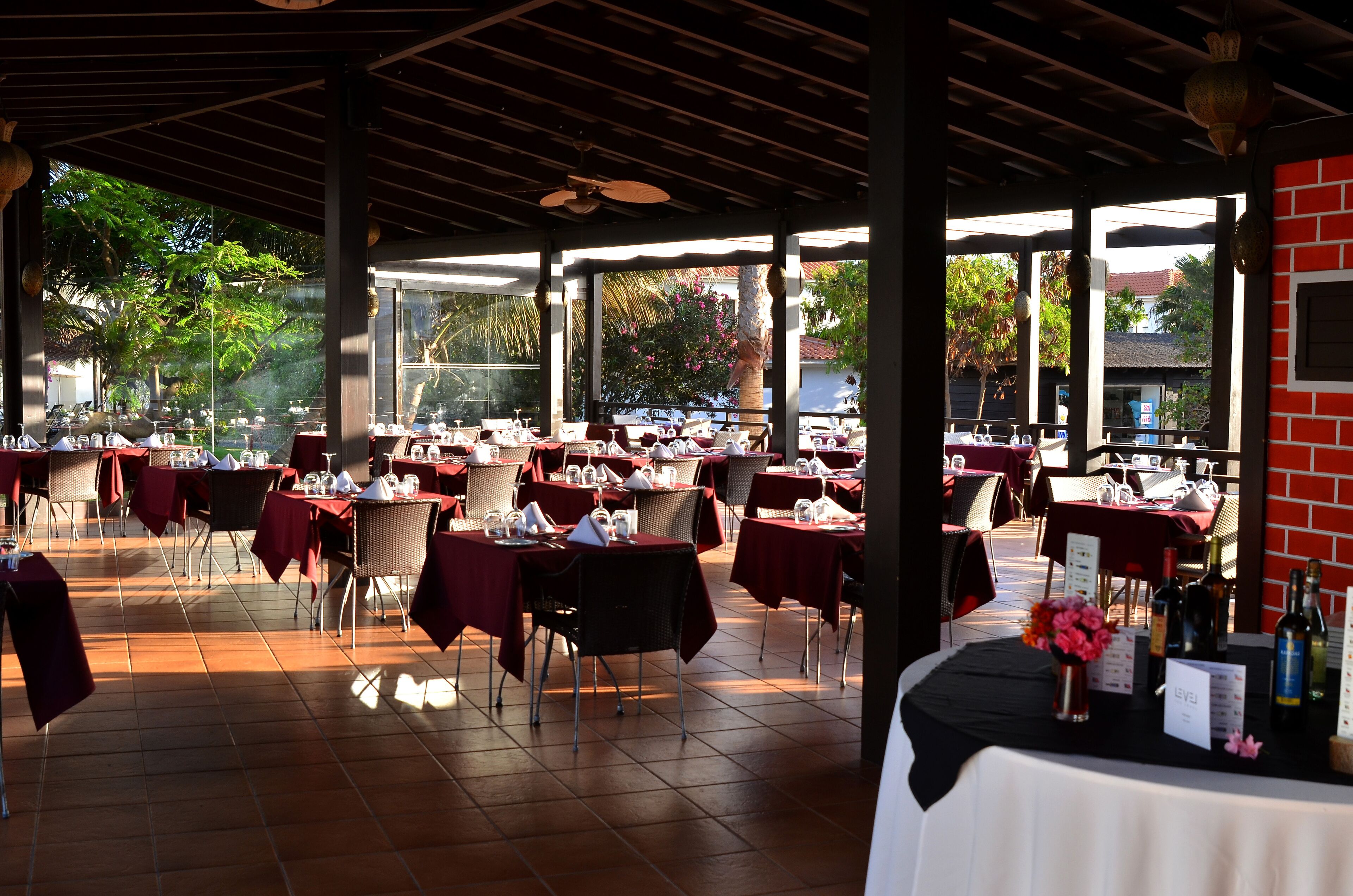 Um restaurante ao ar livre com várias mesas e cadeiras, coberto por um telhado com ventoinha e grandes janelas que oferecem vista da região.