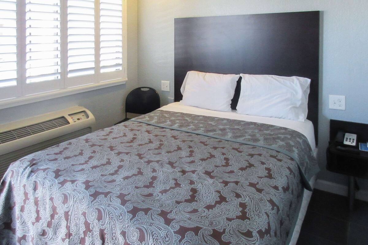 Quarto de hotel com uma cama grande, uma mesa de cabeceira com telefone e uma janela com persianas.