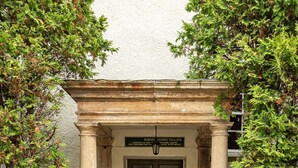 Entrada de pedra com uma placa acima da porta, ladeada por grades de metal verde e cercada por folhagem.