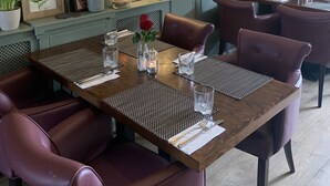 Interior de restaurante com mesa de jantar de madeira, cadeiras estofadas, obras de arte emolduradas nas paredes e um candelabro decorativo.