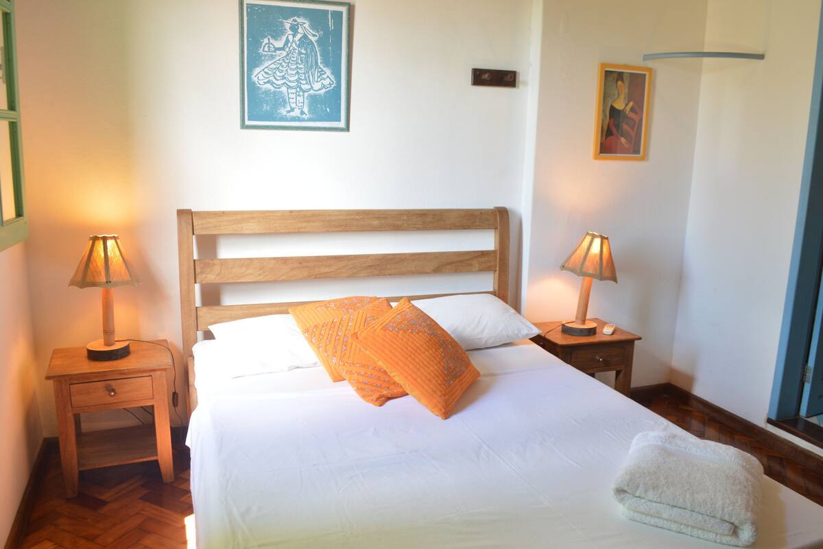 Uma cama bem arrumada com lençóis brancos e um travesseiro laranja, com duas mesas de cabeceira com luminárias, em um quarto com piso de madeira e quadros nas paredes.
