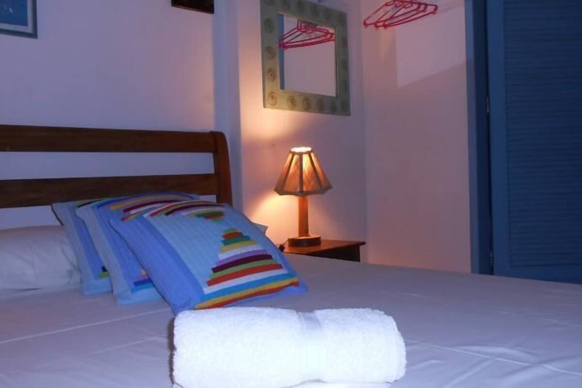Cama bem arrumada com almofada listrada e toalha branca. Tem cabeceira de madeira, abajur e uma porta ao fundo.