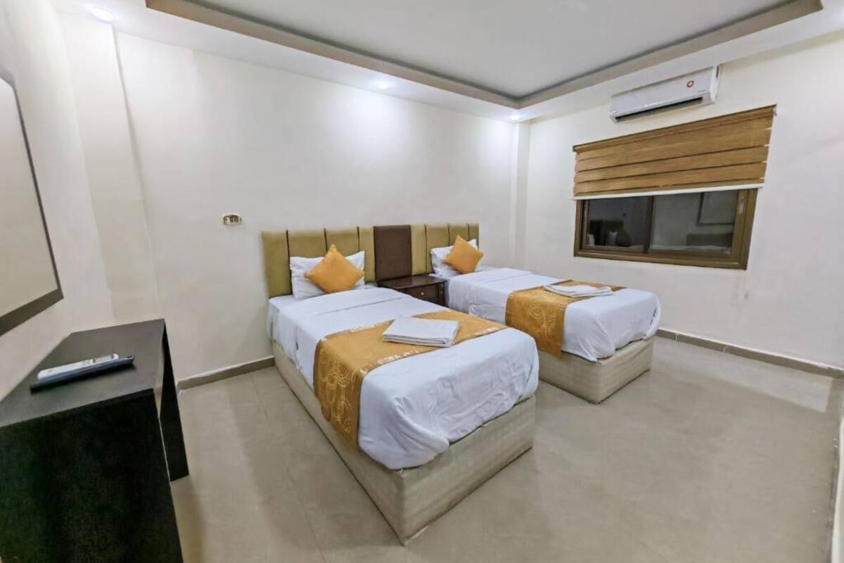 Quarto de hotel com duas camas, uma mesinha de cabeceira, uma janela com persianas e um aparelho de ar condicionado.