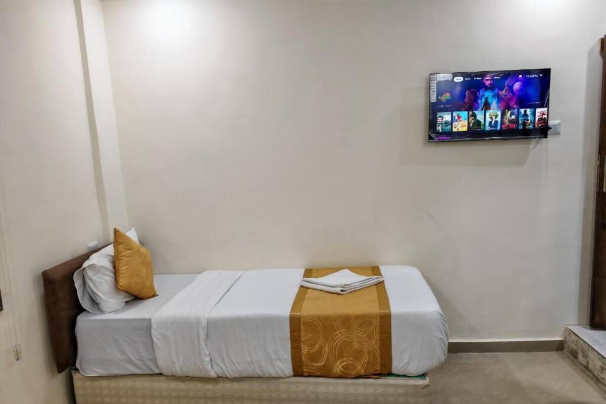 Uma cama de solteiro com colcha branca e marrom, uma televisão na parede exibindo um filme, e uma pequena mesa de cabeceira com um abajur.