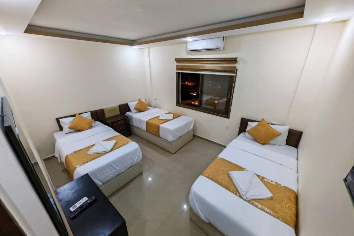 Quarto de hotel com três camas, uma pequena mesa e janela com vista para o exterior.