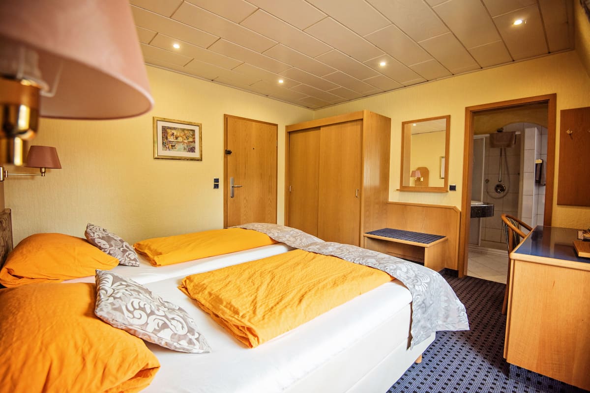 Quarto de hotel com duas camas, um guarda-roupa de madeira, banheiro com chuveiro e uma escrivaninha com cadeira.