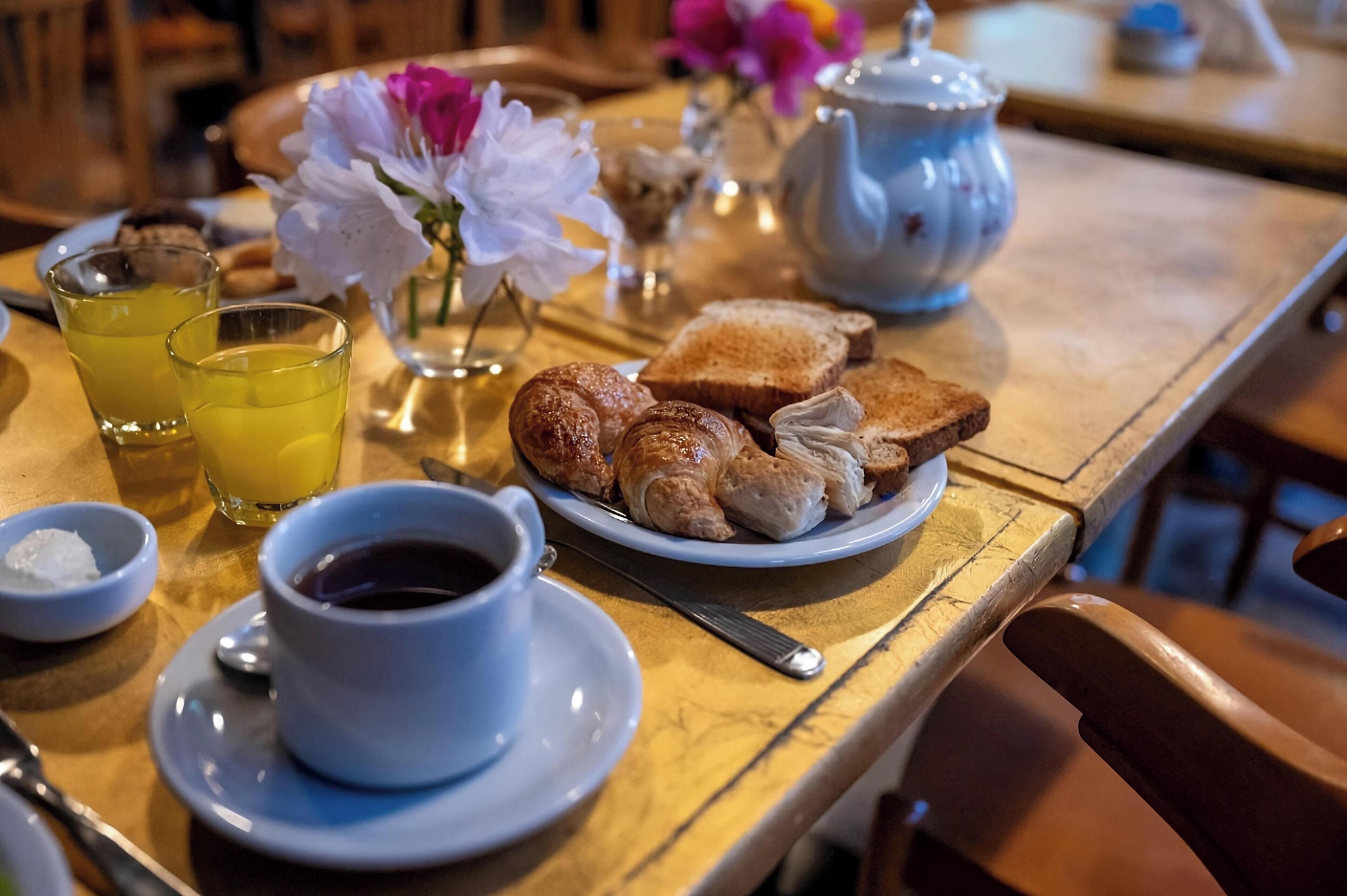 Uma mesa de pequeno-almoço com croissants, torradas, um bule de chá e uma chávena de café.