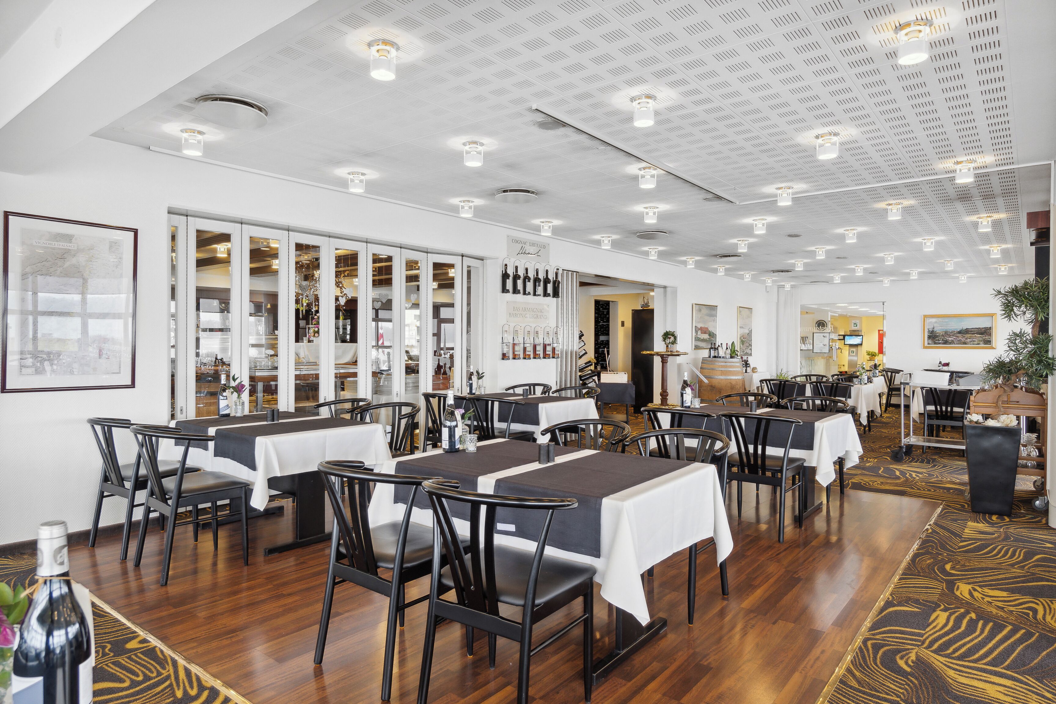 Um restaurante com várias mesas e cadeiras, um tapete estampado e um teto com iluminação embutida.