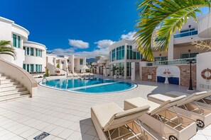 Uma área de piscina em resort, com piscina grande, espreguiçadeiras e escadaria que dá para os andares superiores.