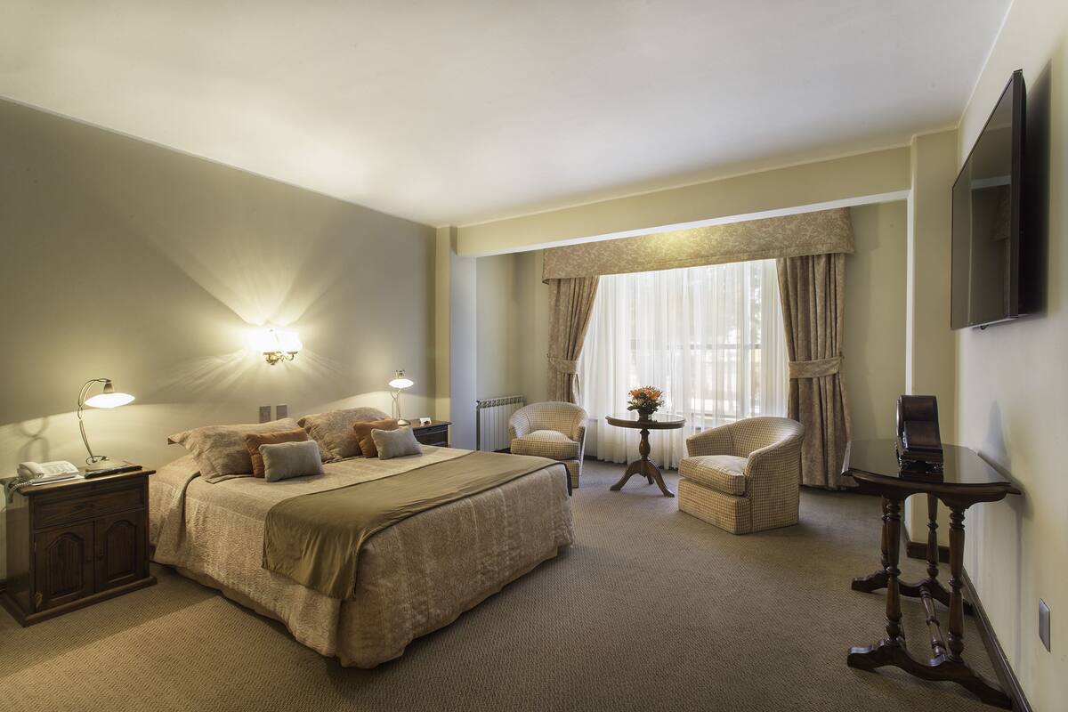 Quarto de hotel com uma cama grande, duas poltronas, uma mesinha, televisão e uma luminária.