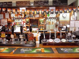 Um balcão de bar com diversas torneiras, incluindo cervejas IPA e Carlsberg, e vários tipos de bebidas alcoólicas.