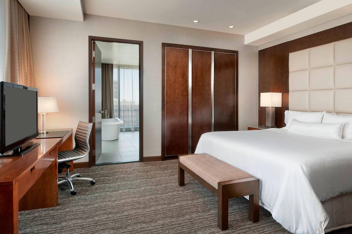 Quarto de hotel com uma cama grande, uma escrivaninha com computador, uma cadeira, uma TV, um banco e um guarda-roupa.
