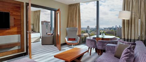 Quarto de hotel com uma grande janela com vista para a cidade, um sofá, uma mesa de centro, uma cadeira e uma pequena mesa com um vaso.