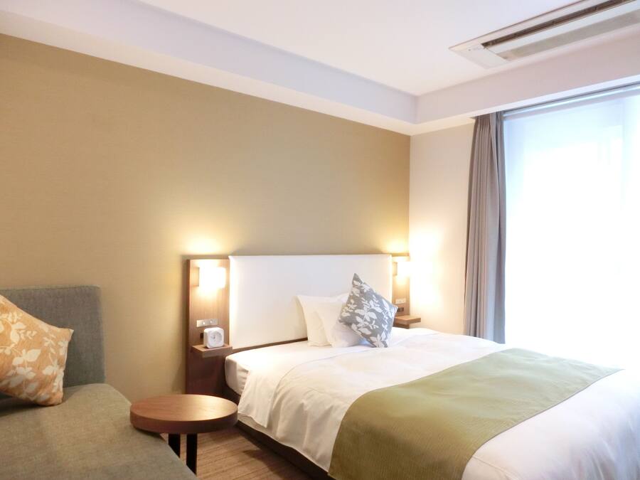 Quarto de hotel com cama, mesa de cabeceira, luminária, cadeira e janela com cortinas.