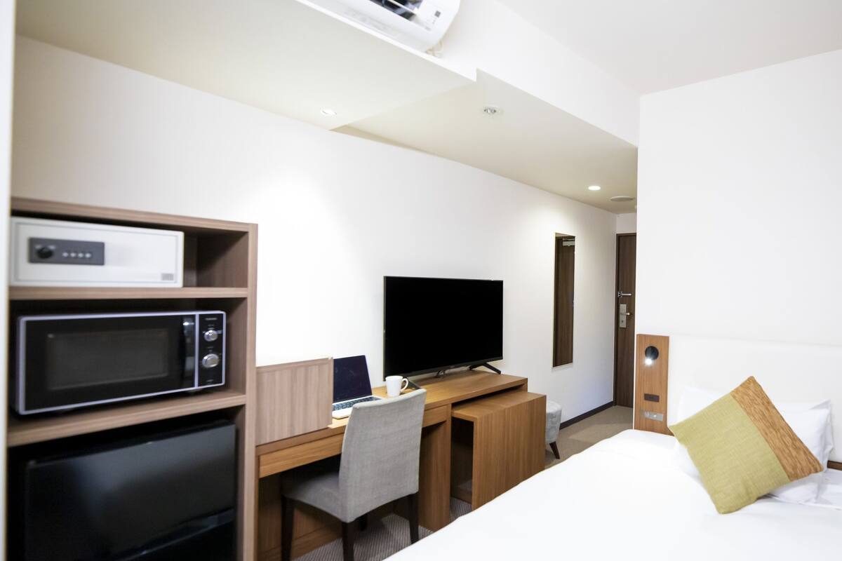 Quarto de hotel com cama, escrivaninha com computador e televisão de tela plana, micro-ondas e pequeno frigorífico.