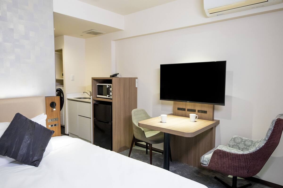 Quarto de hotel com cama, escrivaninha, cadeira, televisão e cozinha compacta.