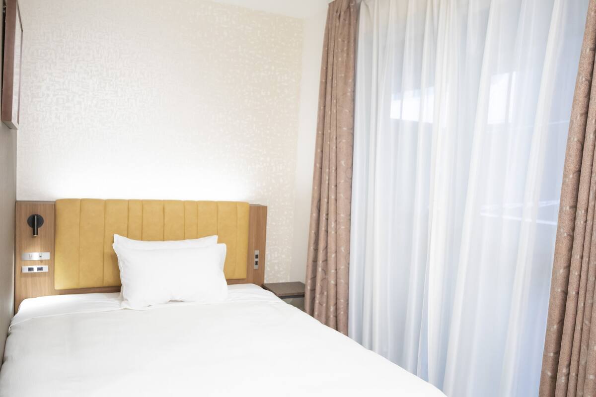 Quarto de hotel com cama, cabeceira, mesa de cabeceira e janela com cortinas.