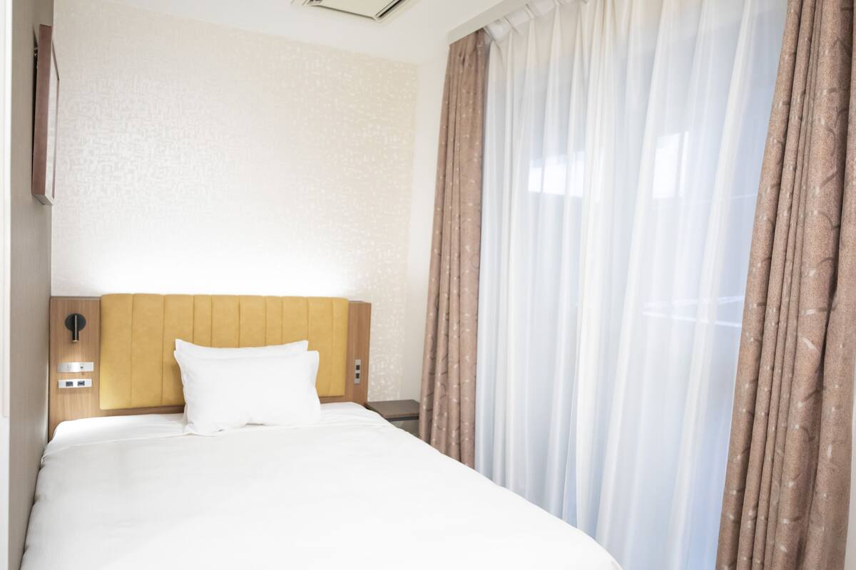 Quarto de hotel com cama, cabeceira, luminária na parede e janela com cortinas.