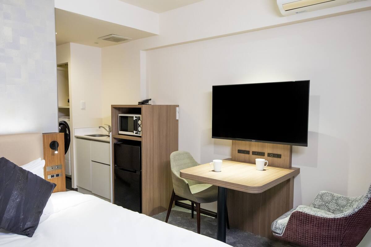Quarto de hotel com uma cama, uma escrivaninha com duas chávenas, uma televisão, um micro-ondas e uma cadeira.