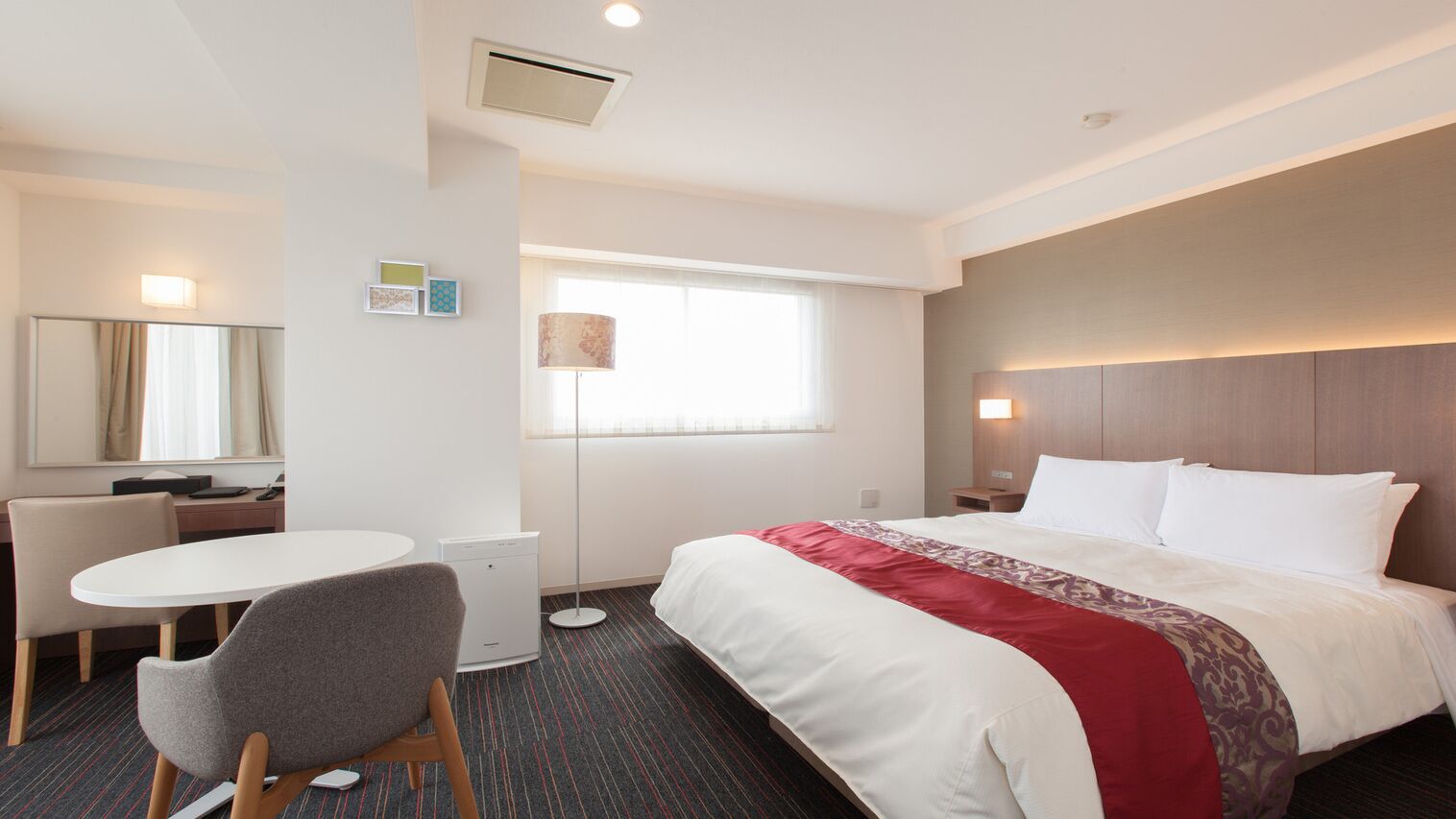 Quarto de hotel com uma cama, uma escrivaninha, uma cadeira, um abajur e uma janela.