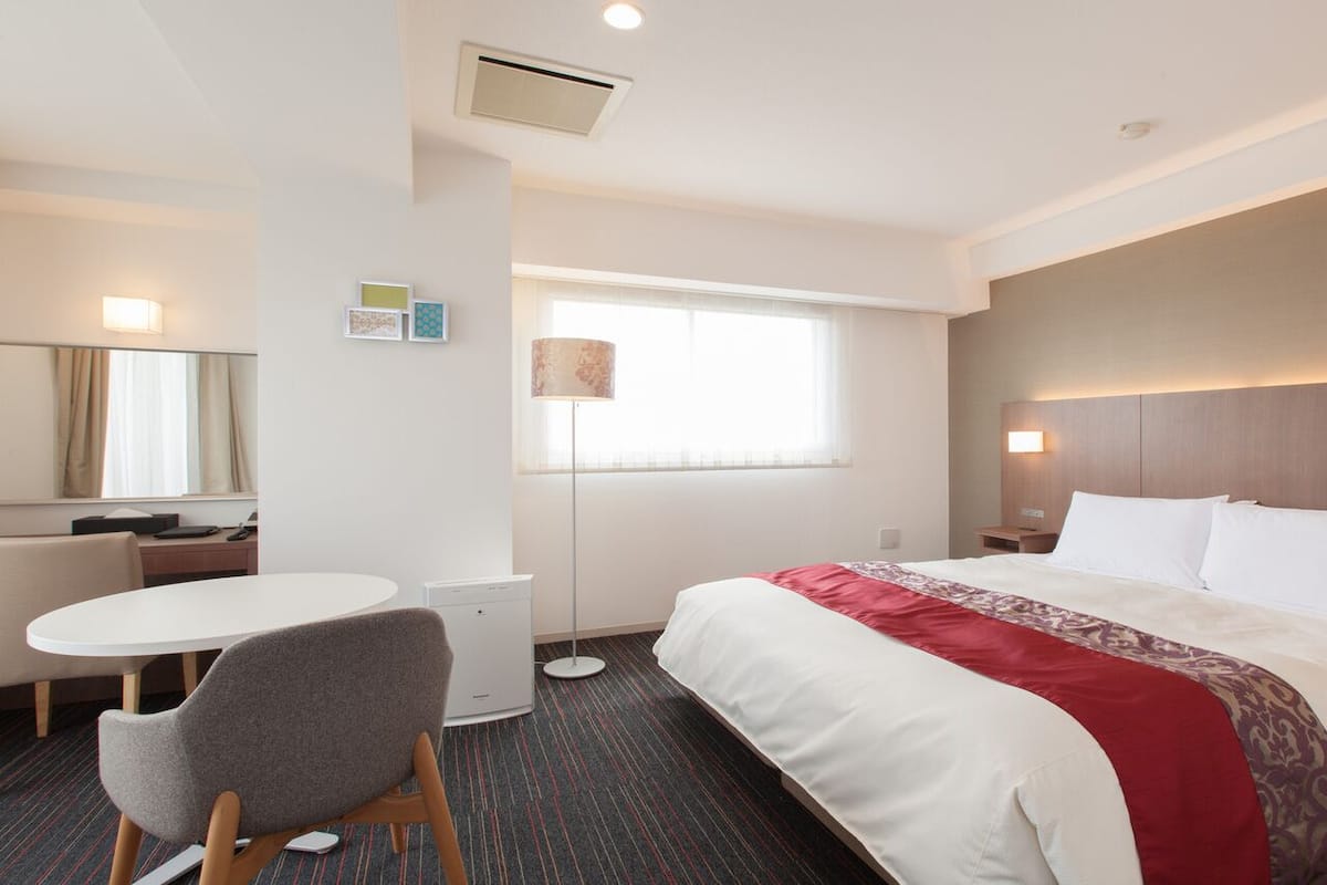 Quarto de hotel com uma cama, uma escrivaninha, uma cadeira, um abajur e uma janela.