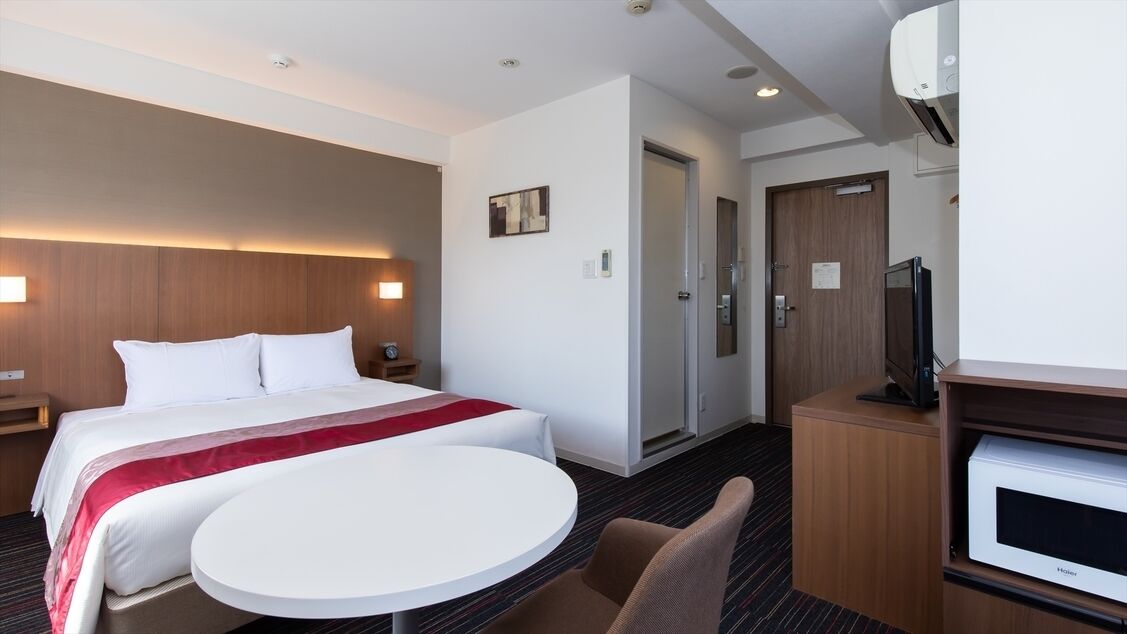 Quarto de hotel com cama, mesa de cabeceira, luminária, TV e uma pequena mesa redonda com uma cadeira.