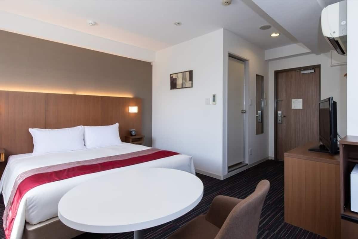 Quarto de hotel com cama, mesa de cabeceira, luminária, TV e uma pequena mesa redonda com uma cadeira.