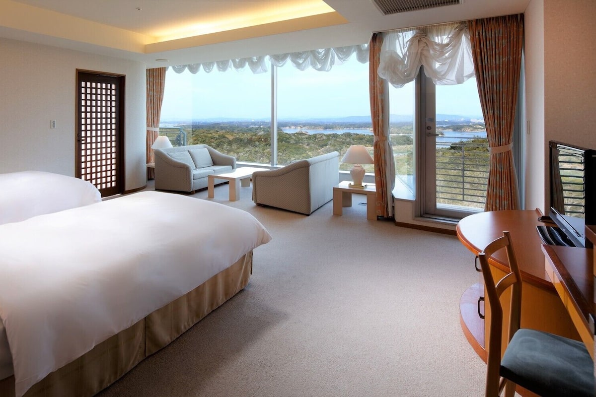 Quarto de hotel com uma cama grande, um sofá, uma escrivaninha e vista para o exterior.