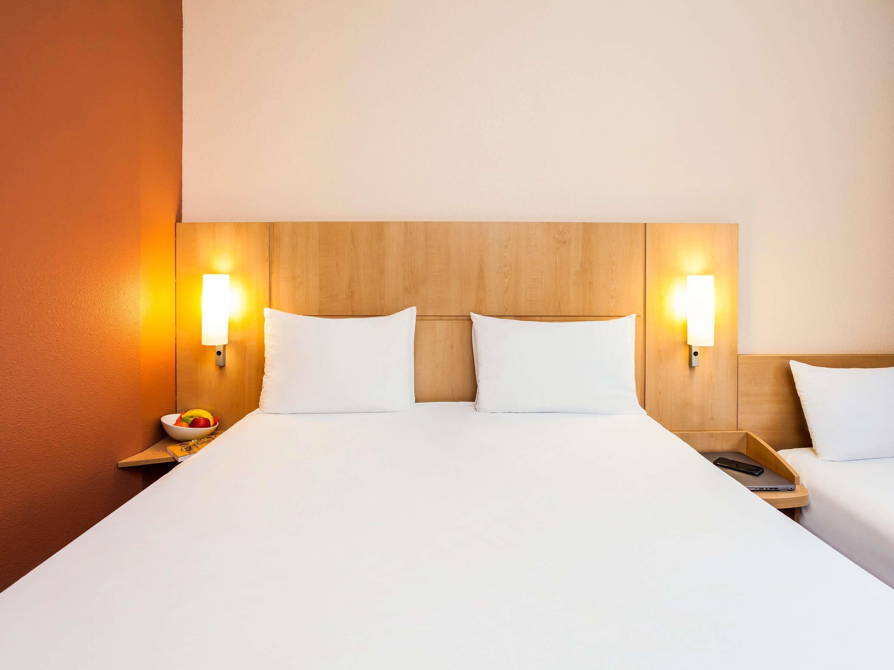 Quarto de hotel com cabeceira de madeira, duas almofadas brancas, edredom branco e duas mesas de cabeceira com abajures.