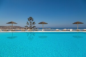 Piscina azul límpida com quatro guarda-sóis, uma palmeira e vista para o mar e colinas ao longe.